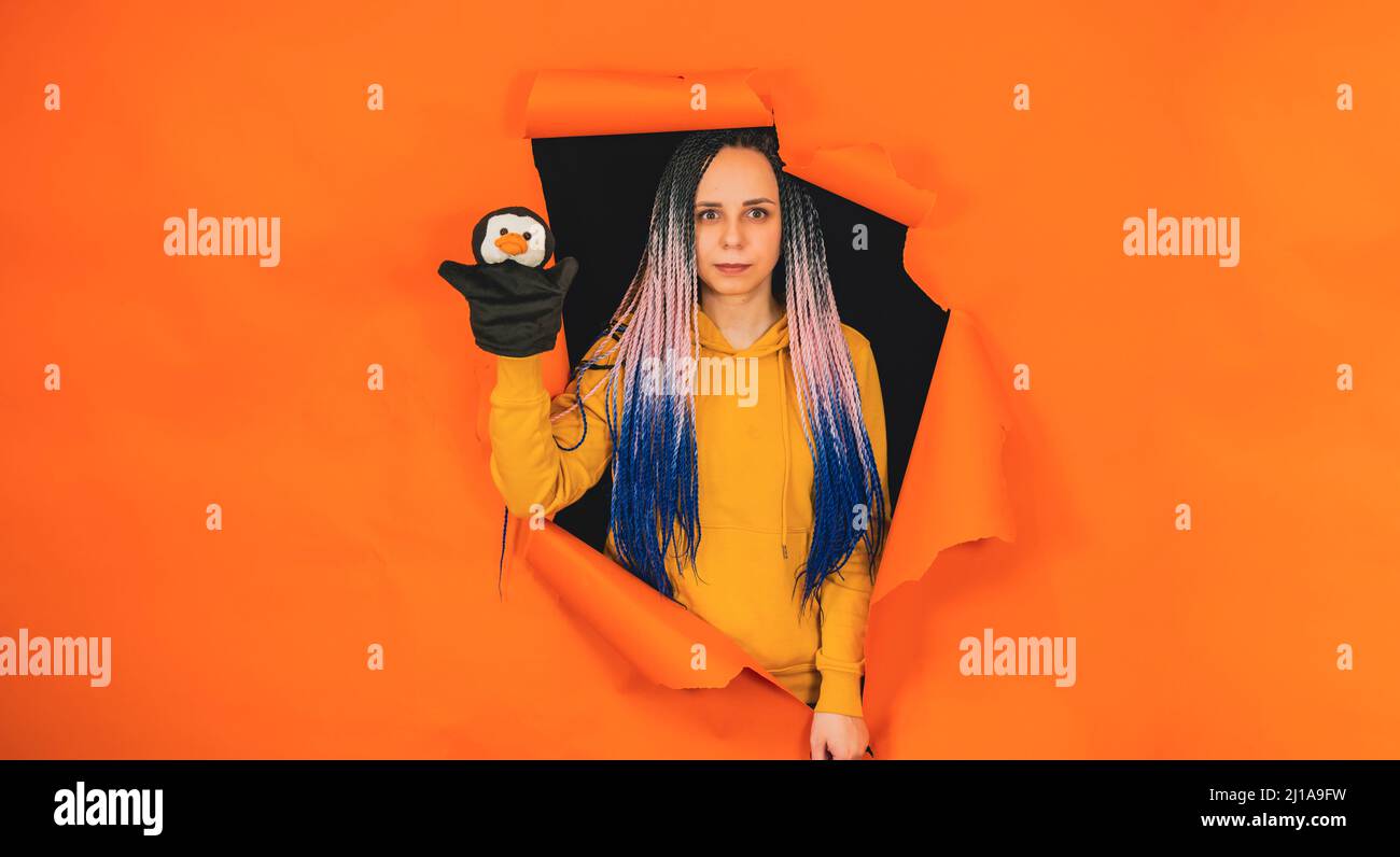 Young woman with soft puppet toy on hand looking out of hole of orange ...