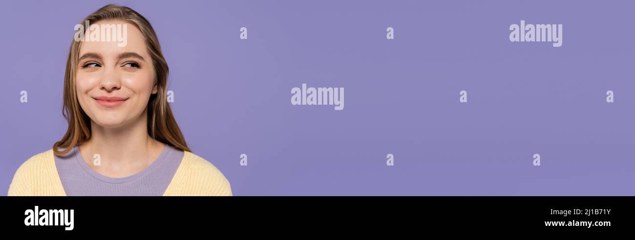 sly young woman looking away and smiling isolated on purple, banner ...