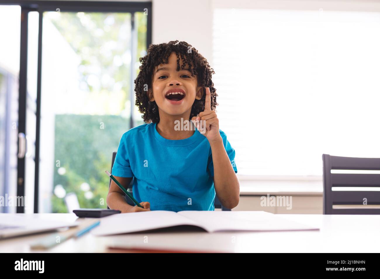 Portrait of cheerful hispanic boy with arm raised studying at table ...