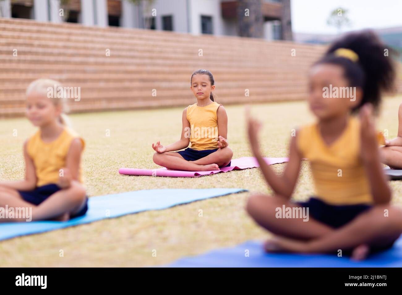 Multiracial elementary schoolgirls meditating while sitting on yoga mat ...