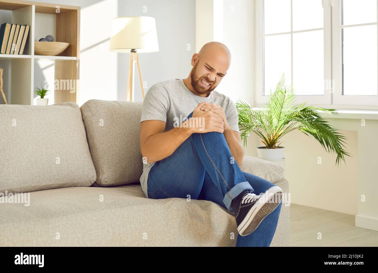 Young man sitting on sofa, suffering from knee pain and touching his ...