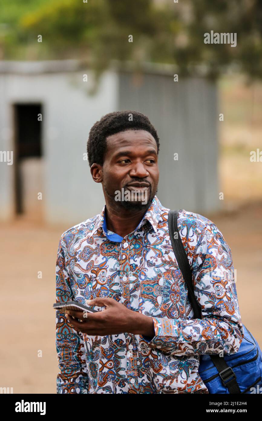 A young Black teacher in a colorful shirt with a phone on his hand ...