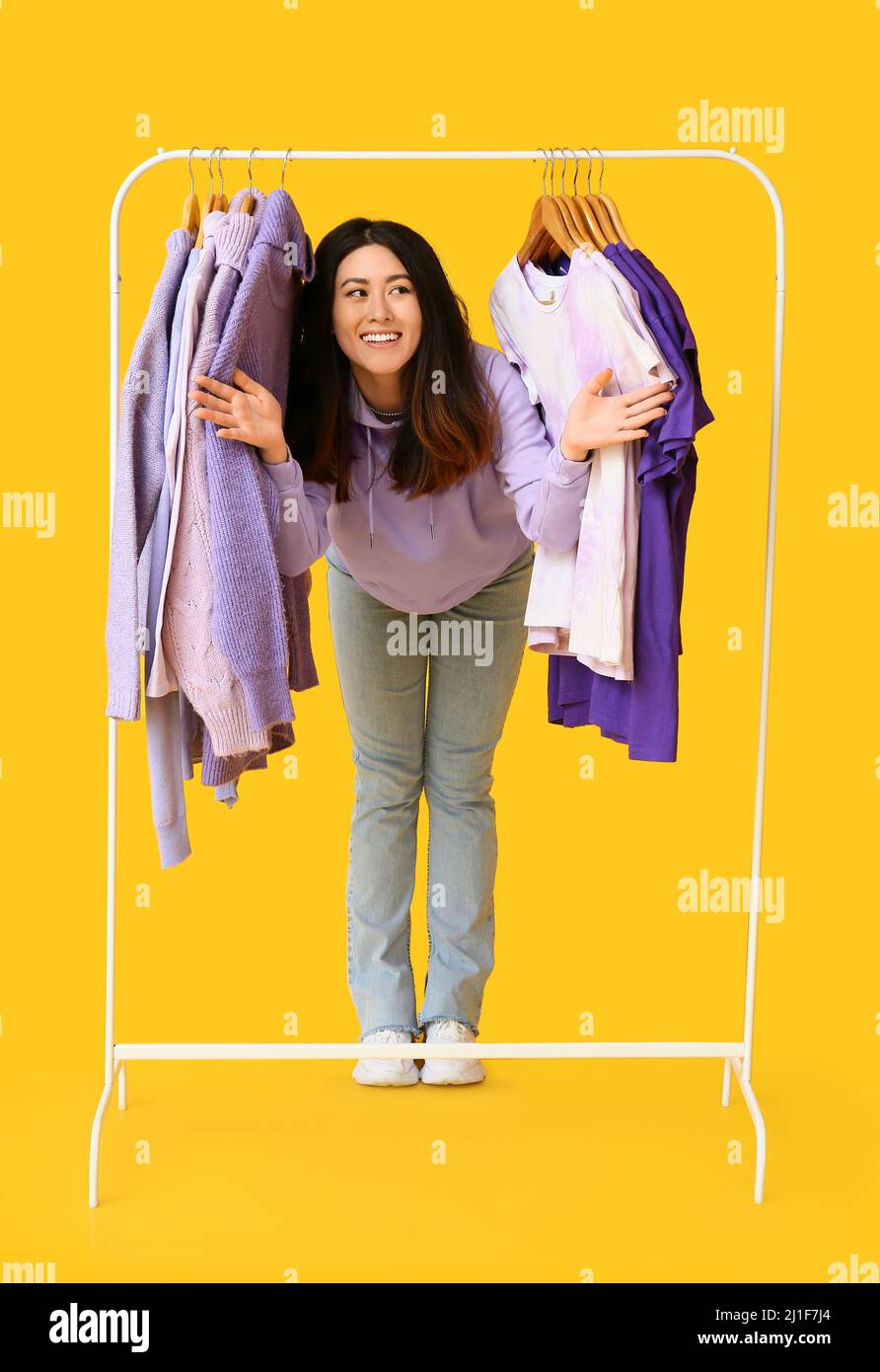 Young Asian woman and rack with clothes in purple shades on yellow ...