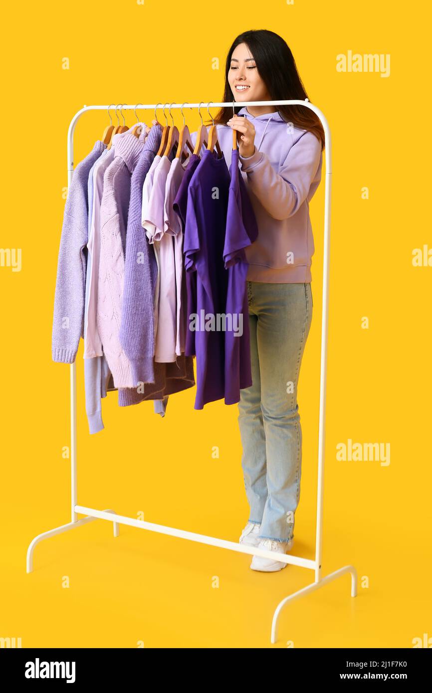 Young Asian woman and rack with clothes in purple shades on yellow ...