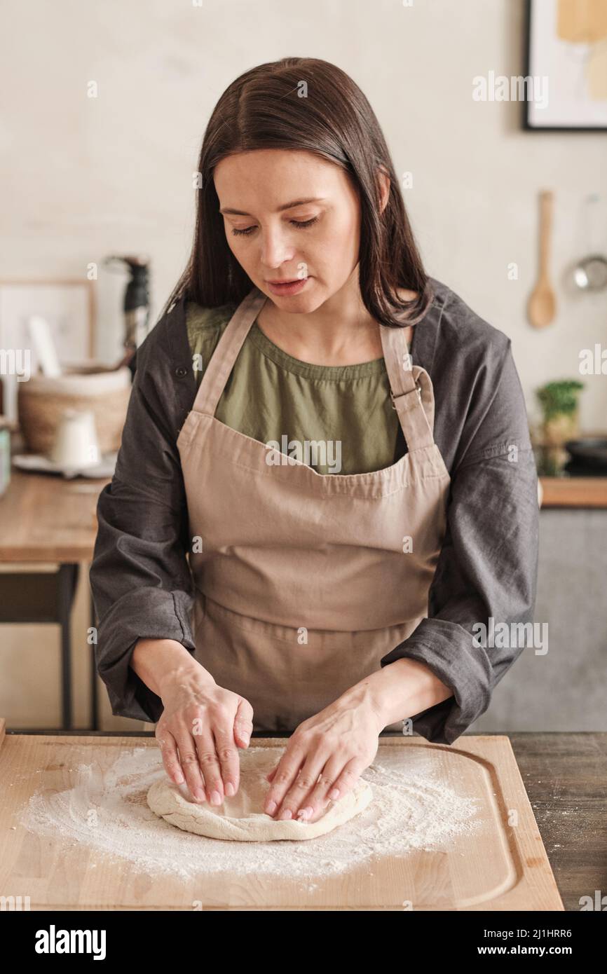 Serious young woman in apron standing at table with wooden board and ...