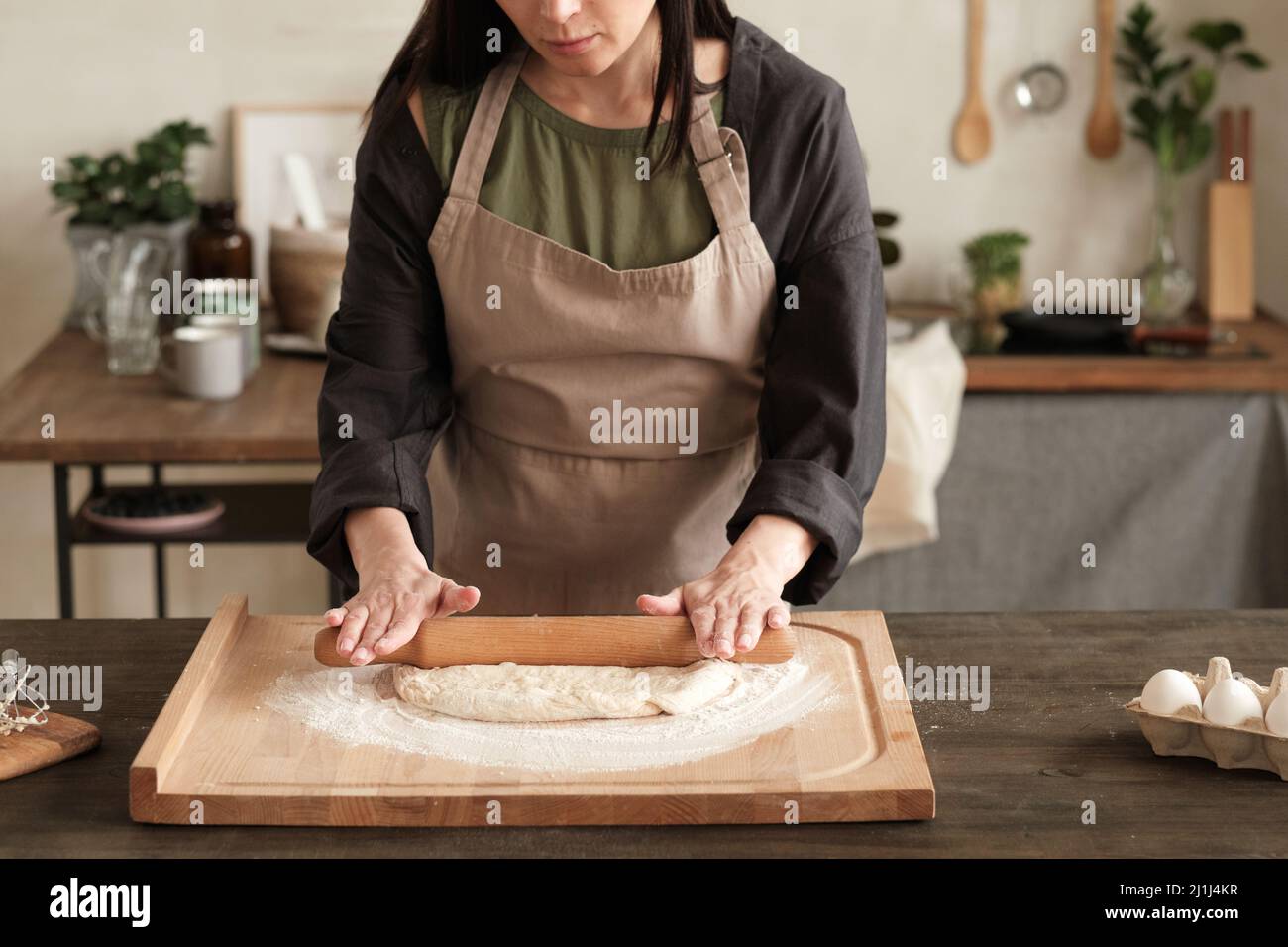 Busy woman in apron standing at table and rolling dough with pin on ...