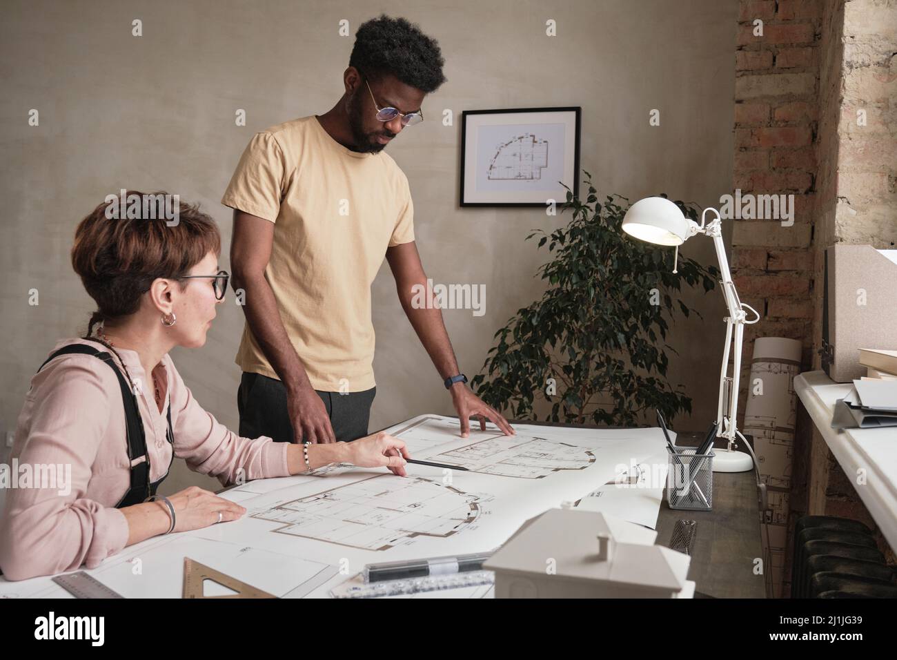 Mature woman in glasses sitting at table and pointing at blueprint while explaining design of ...
