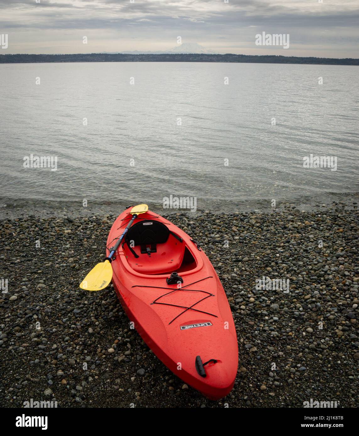 A bright red kayak with a yellow paddle on the rocky shore of Puget ...