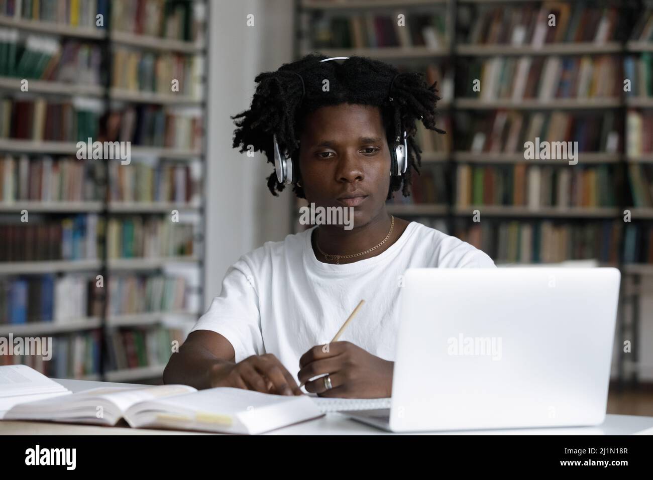 Serious African student guy in headphones makes assignment in library ...
