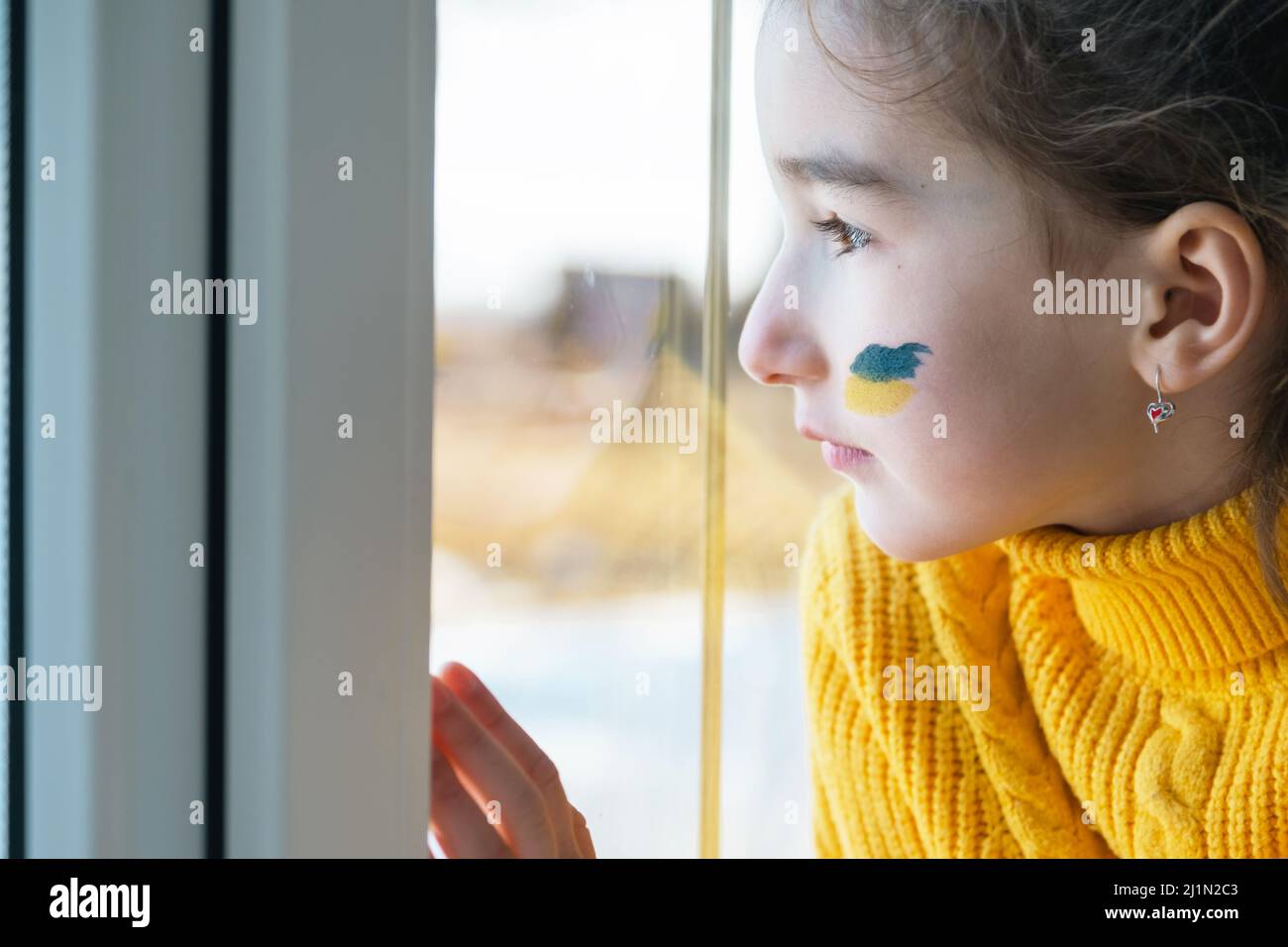 A sad child look at the window with the flag of Ukraine painted on the ...