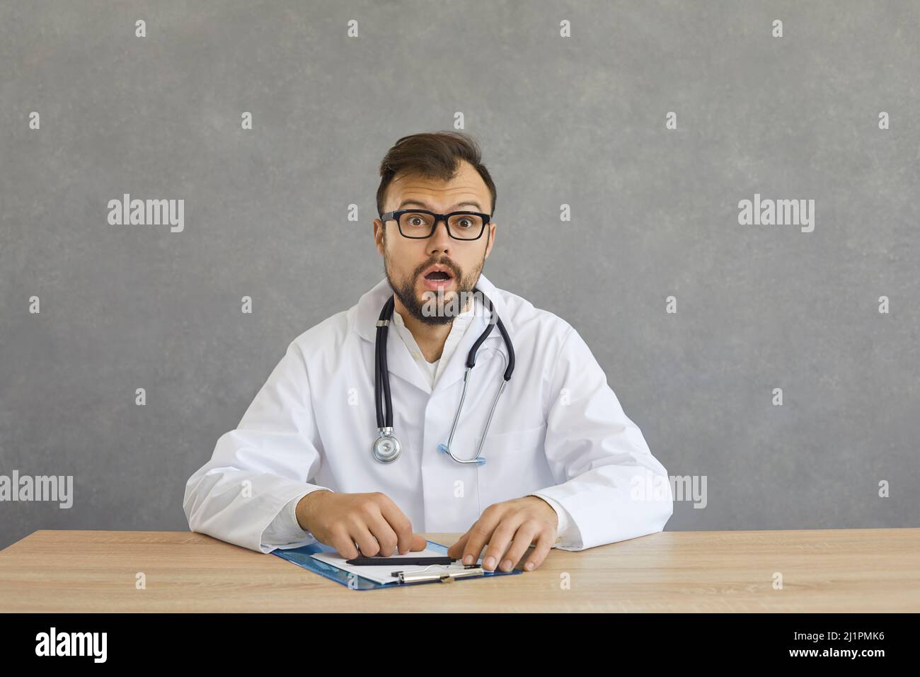 Shocked doctor sitting at desk table looking at camera studio portrait ...