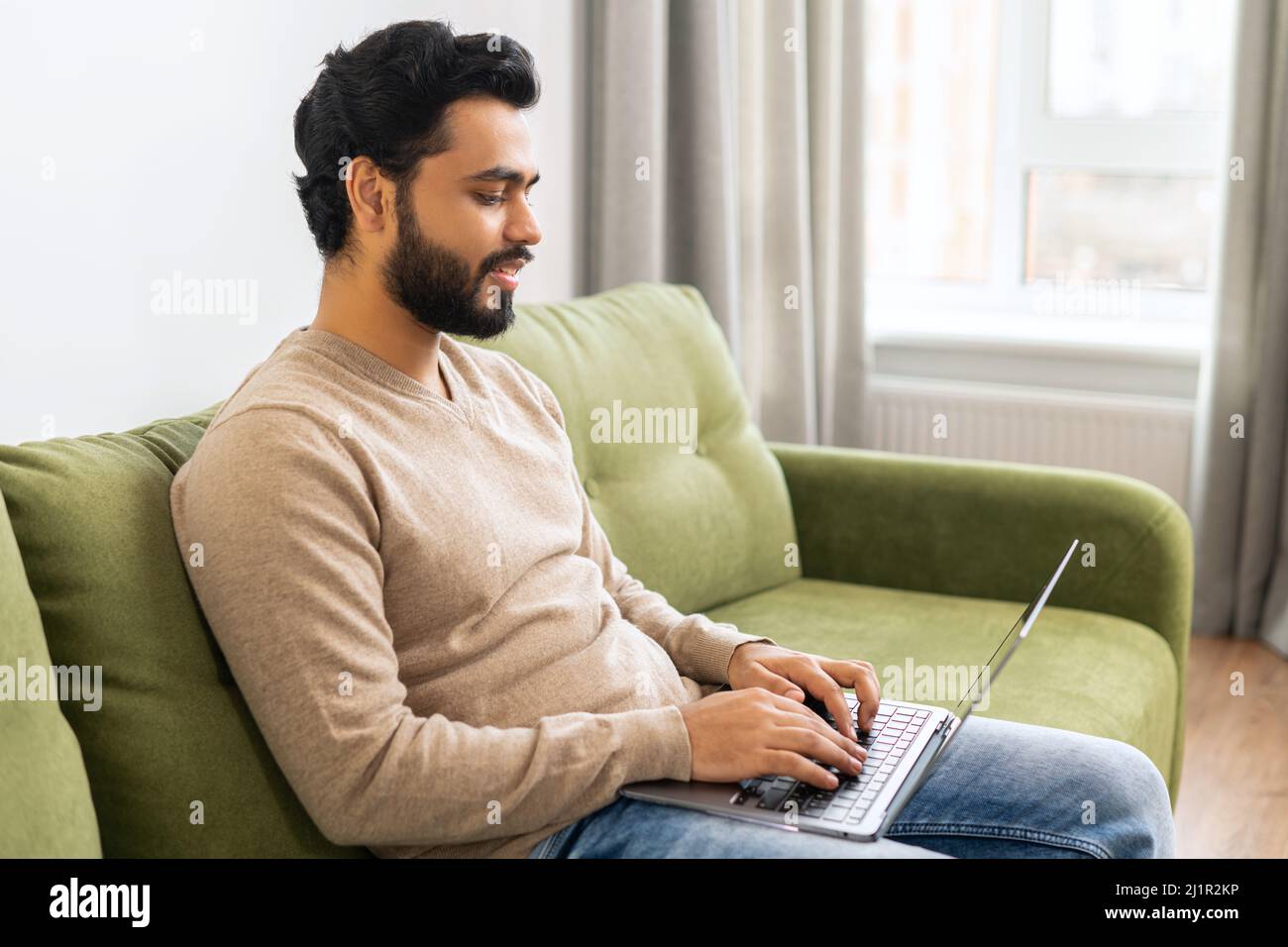 Optimistic ethnic man in casual wear using laptop sitting on the couch ...