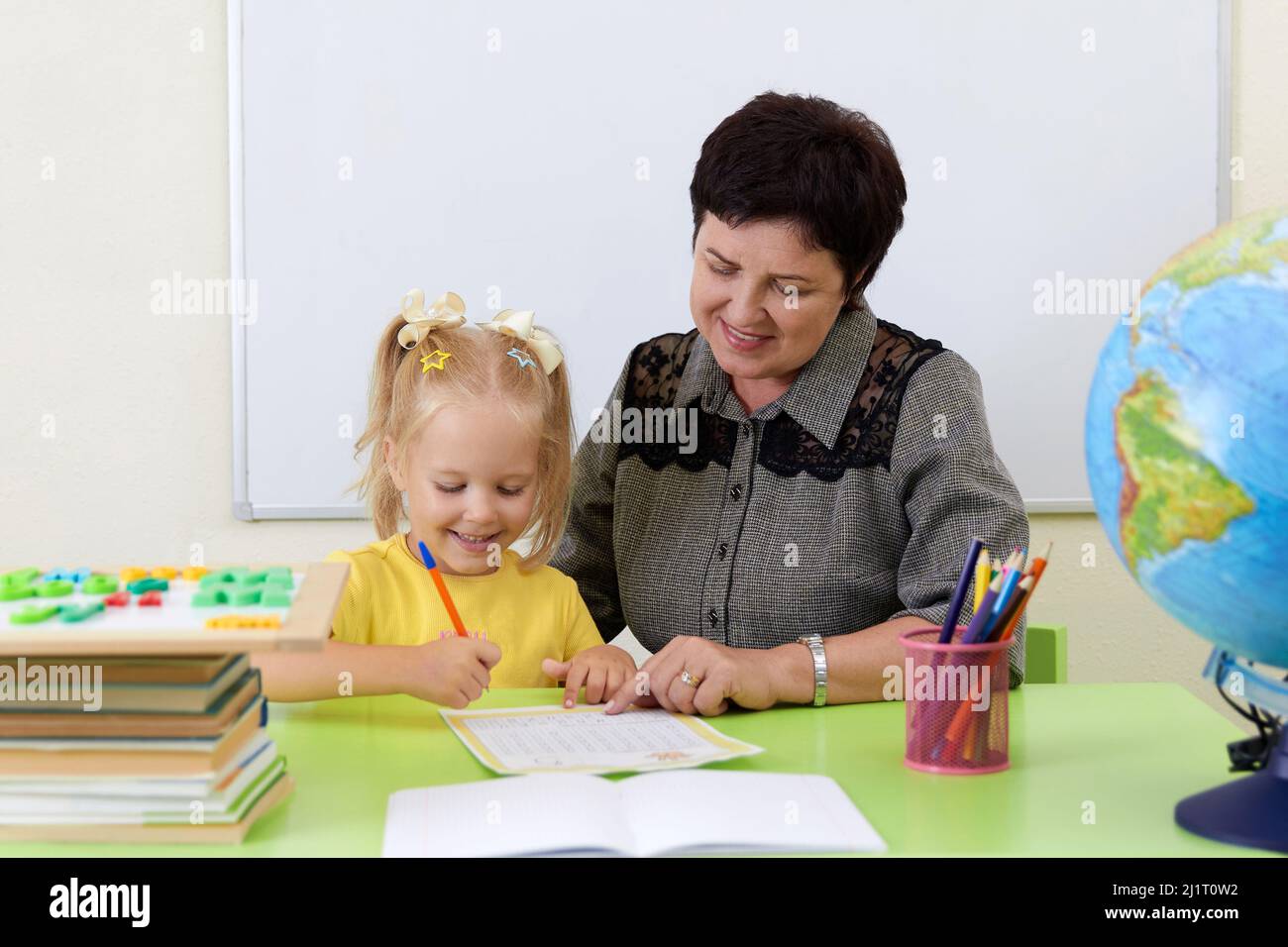 Mature teacher sitting at table teaching little girl how to write Stock ...