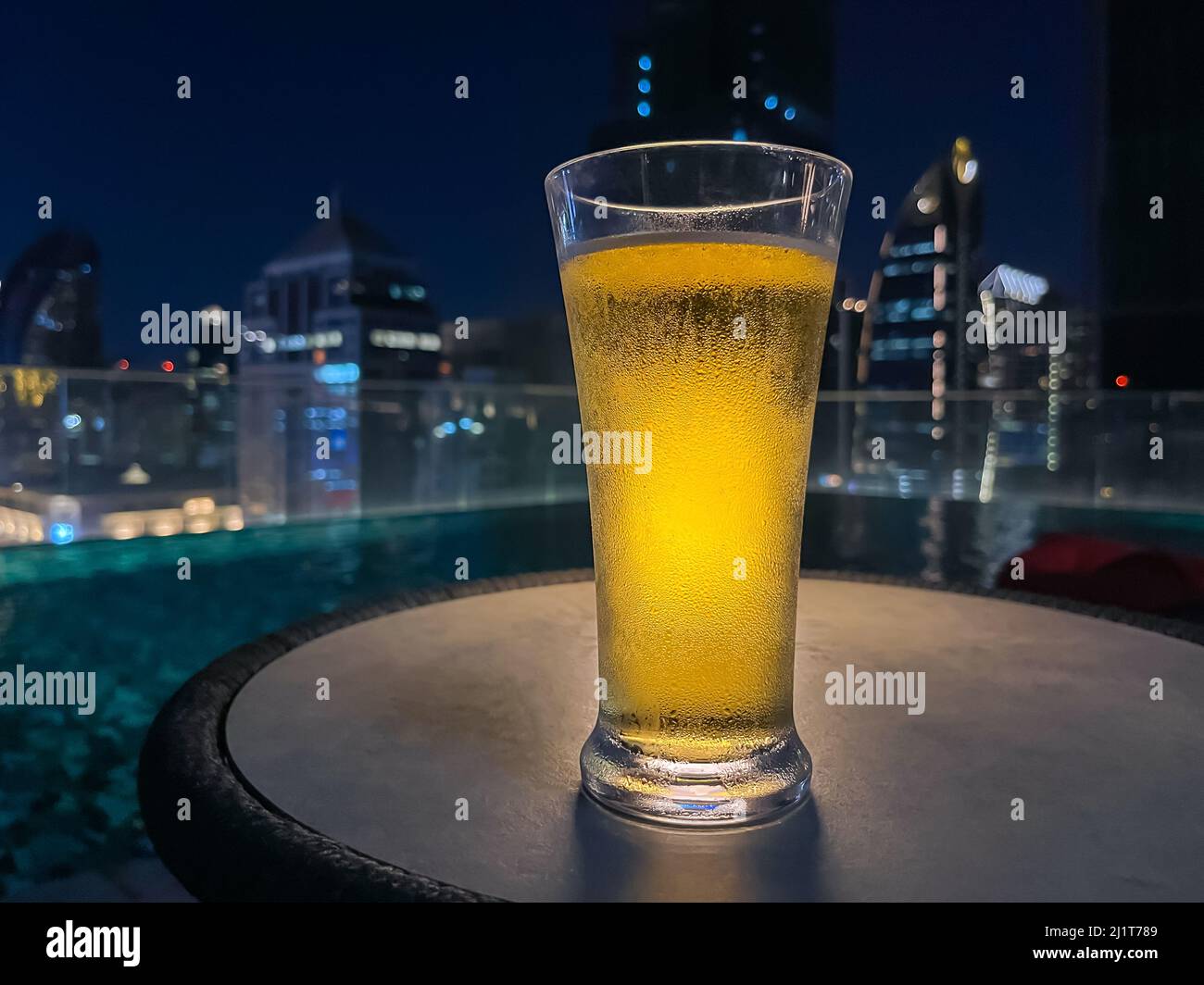 A glass of beer puts on table at rooftop bar with colorful city ...