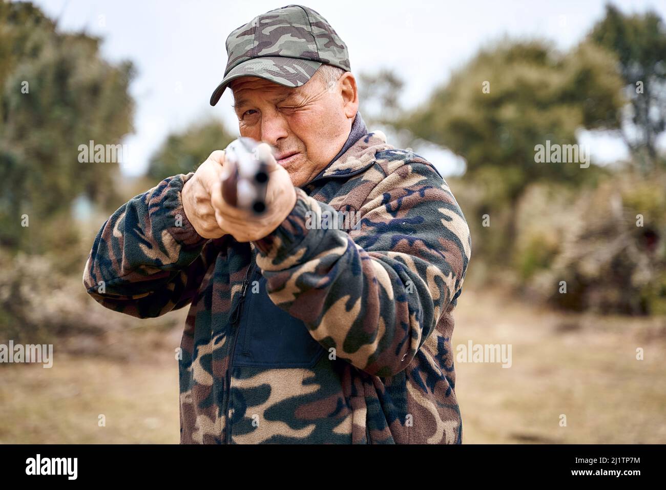 Older man in camouflage clothing and cap squinting and pointing shotgun ...