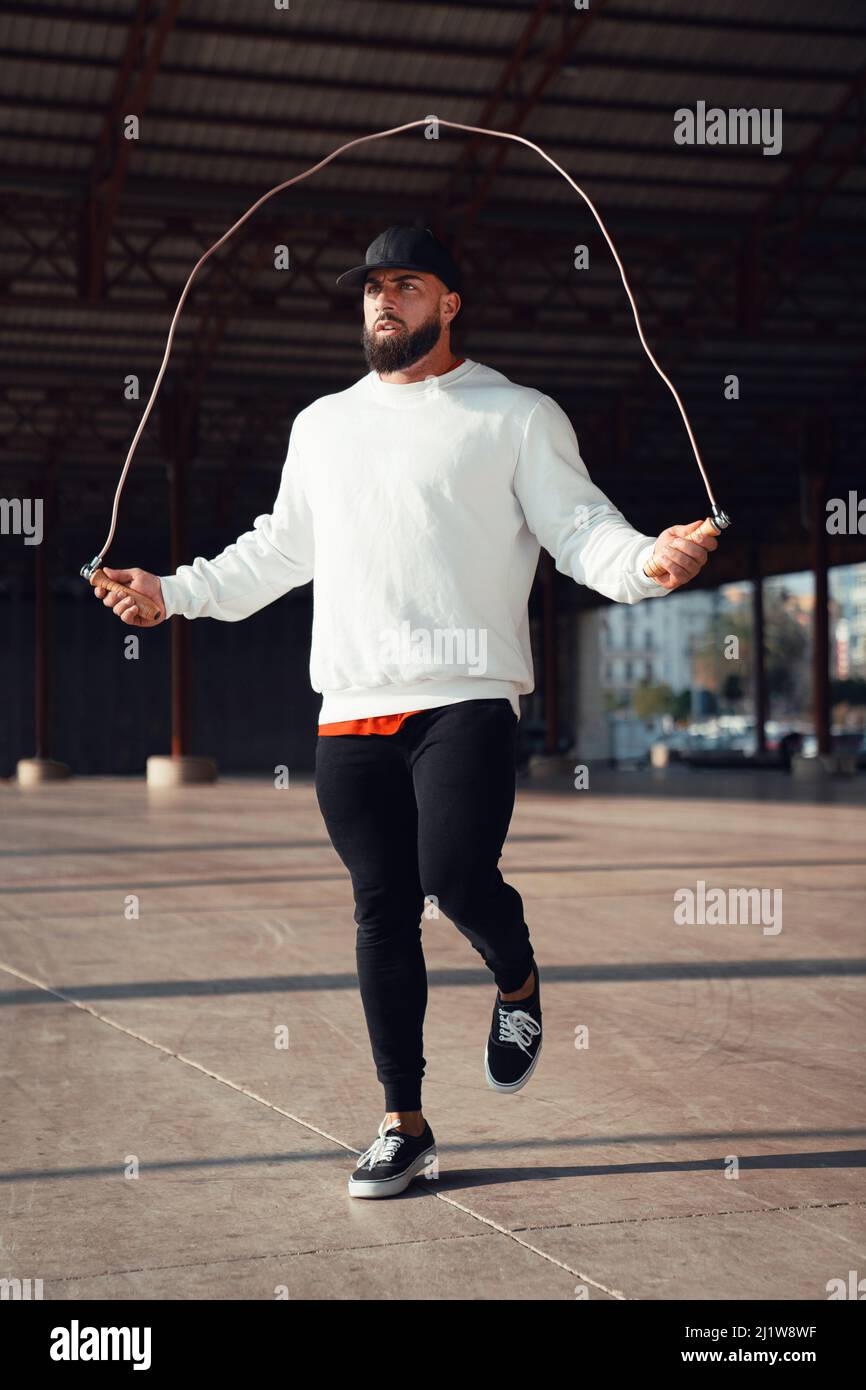 Full body of active bearded male in black cap jumping rope while ...
