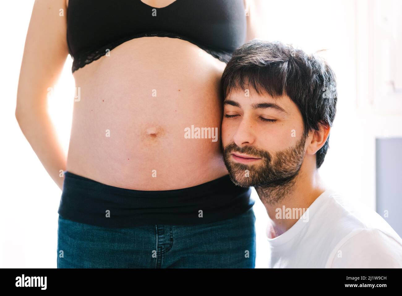 Dreamy Husband With Closed Eyes Touching And Listening To Tummy Of Crop Pregnant Wife At Home