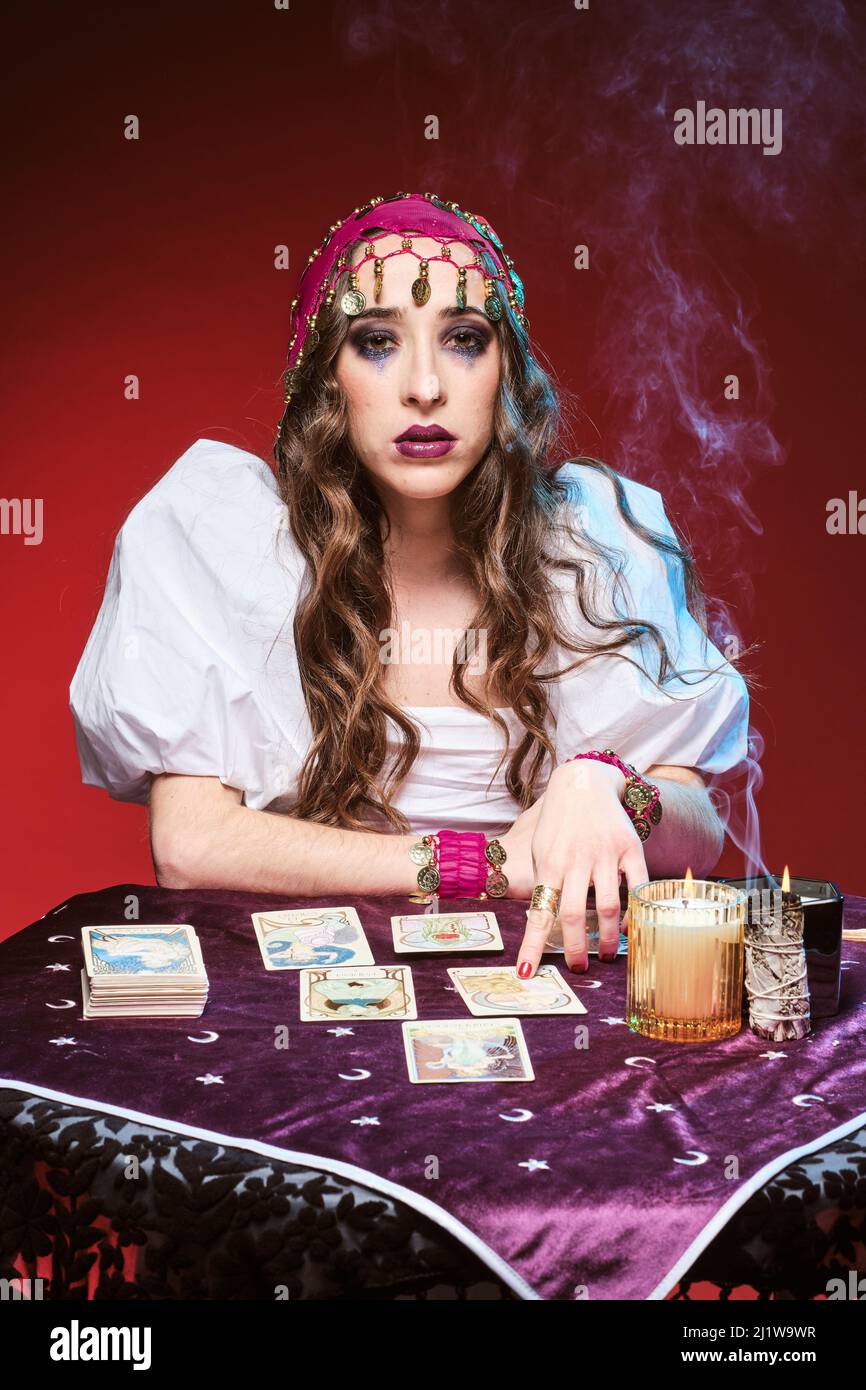 Attractive female soothsayer with makeup sitting at table with tarot cards against burning ...