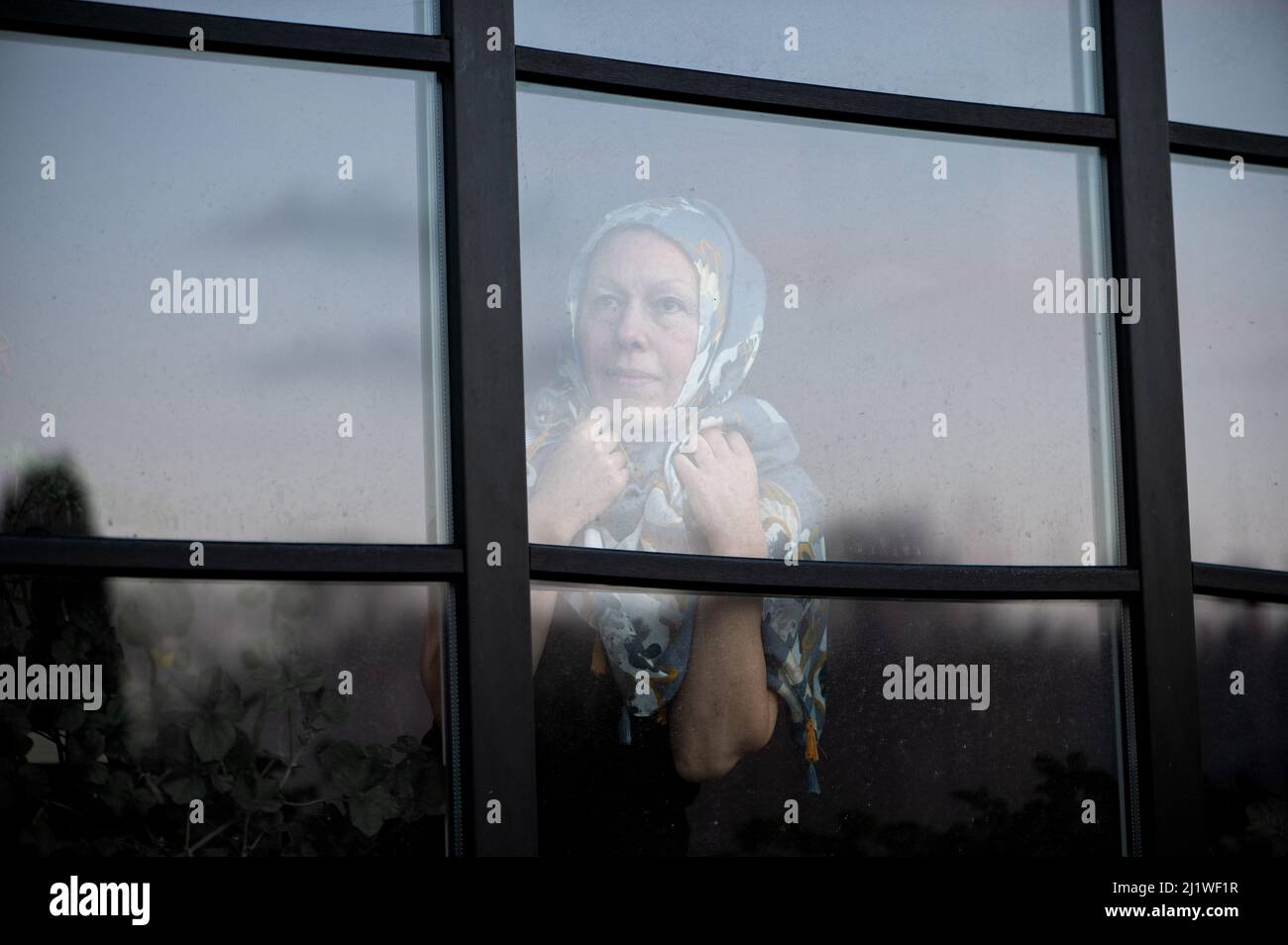 Sad mature woman with scraft on head standing behind dirty window glass ...