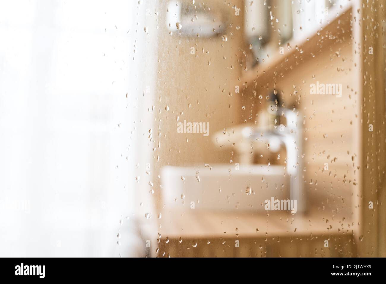 Wet glass shower door in hotel bathroom with blured window, sink and ...