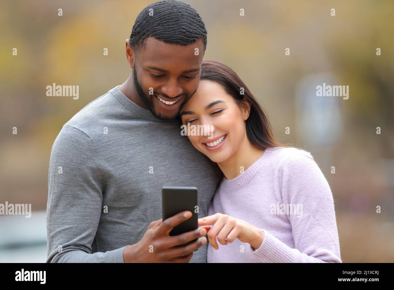 Front view of a happy interracial couple checking smart phone outdoors ...
