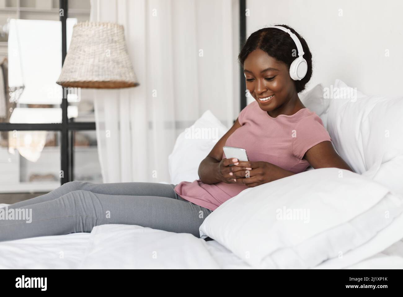 African American Lady Using Mobile Phone Wearing Heaphones In Bedroom ...
