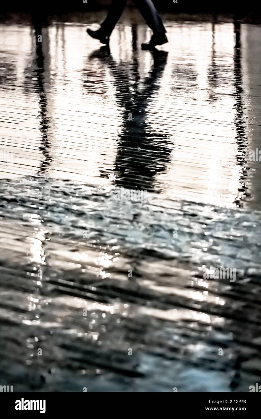 People walk along a rain soaked street made of tiles reflected in the ...