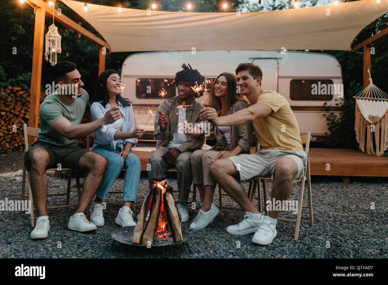 Millennial diverse friends sitting near bonfire with lit sparklers ...