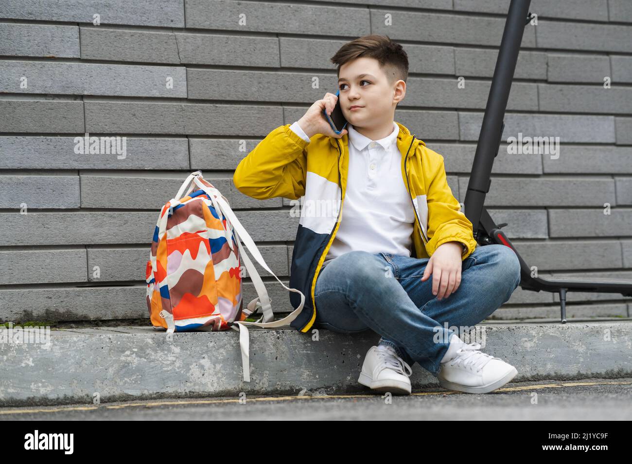 Cute boy talking on the phone while sitting on the sidewalk near his ...