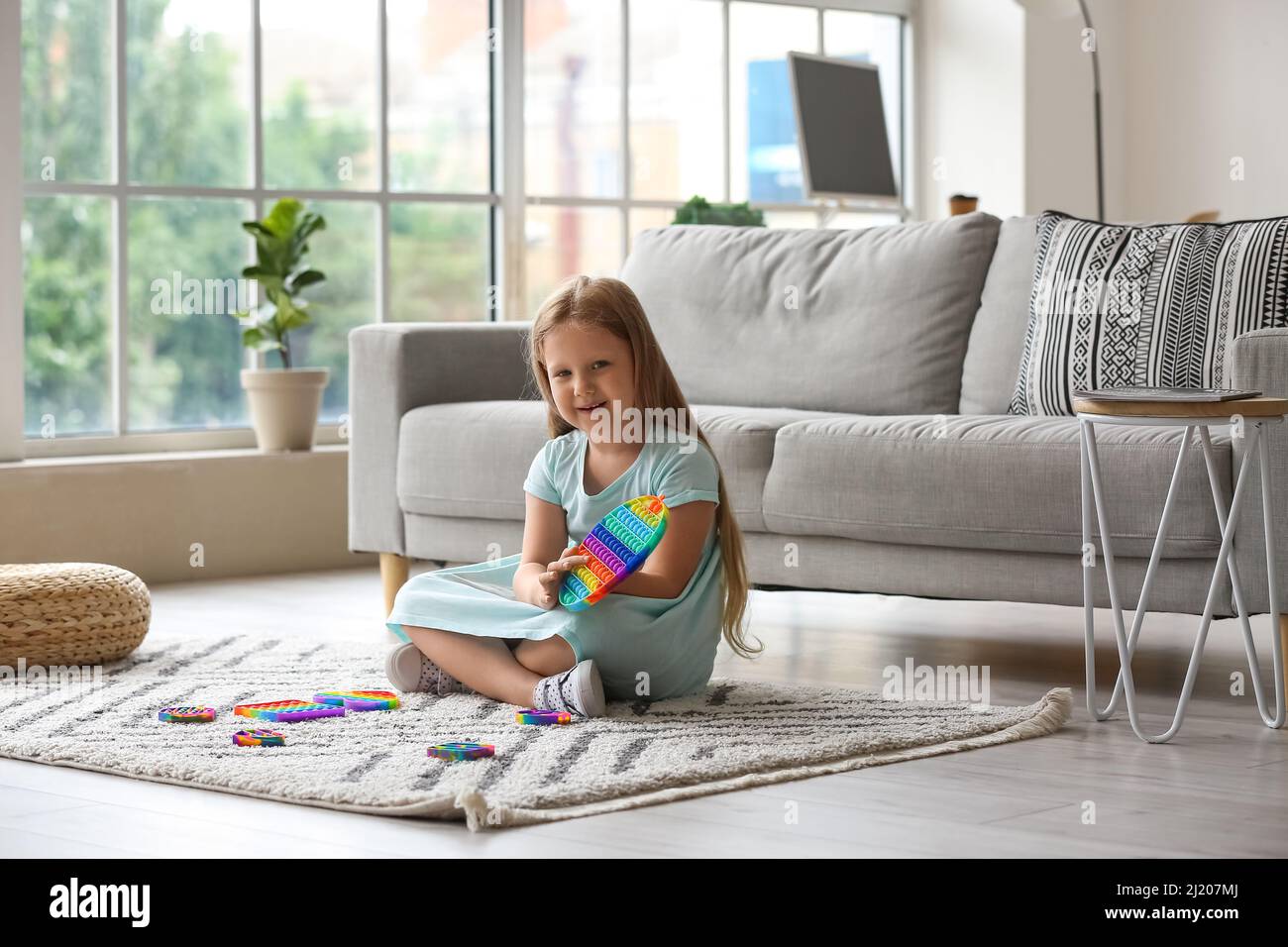 Pretty girl sitting on floor and holding pop it fidget toy in room ...