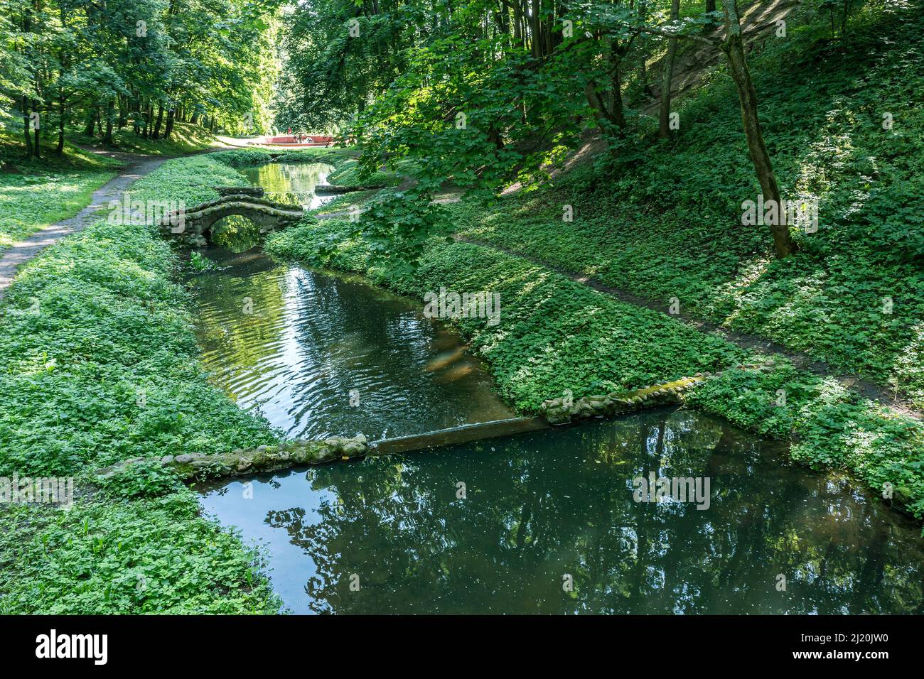 summer park landscape with small arched stone footbridge over creek ...