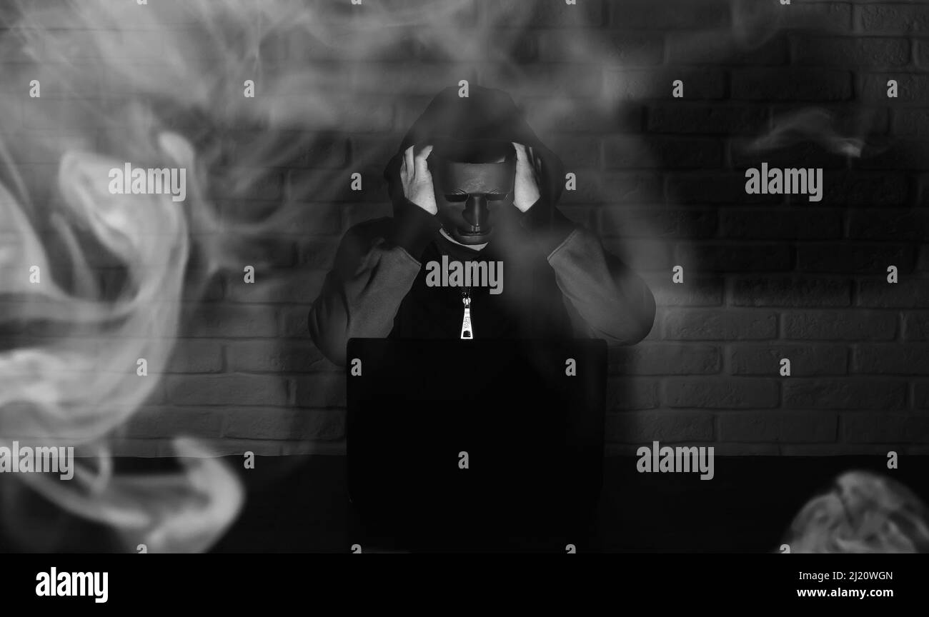 Hacker in black mask and hood at the table in front of the monitor ...