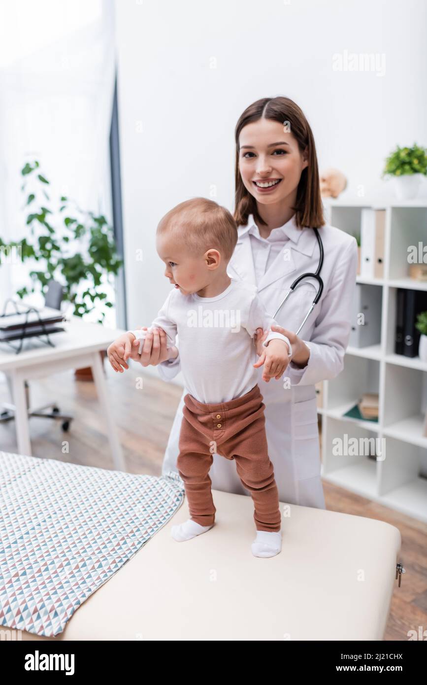 Smiling family doctor holding hands of baby boy on medical couch in ...