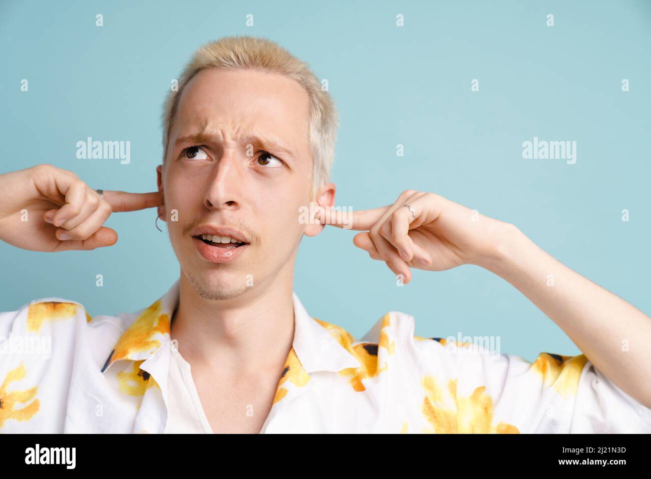Young blonde man in shirt frowning and plugging his ears isolated over ...