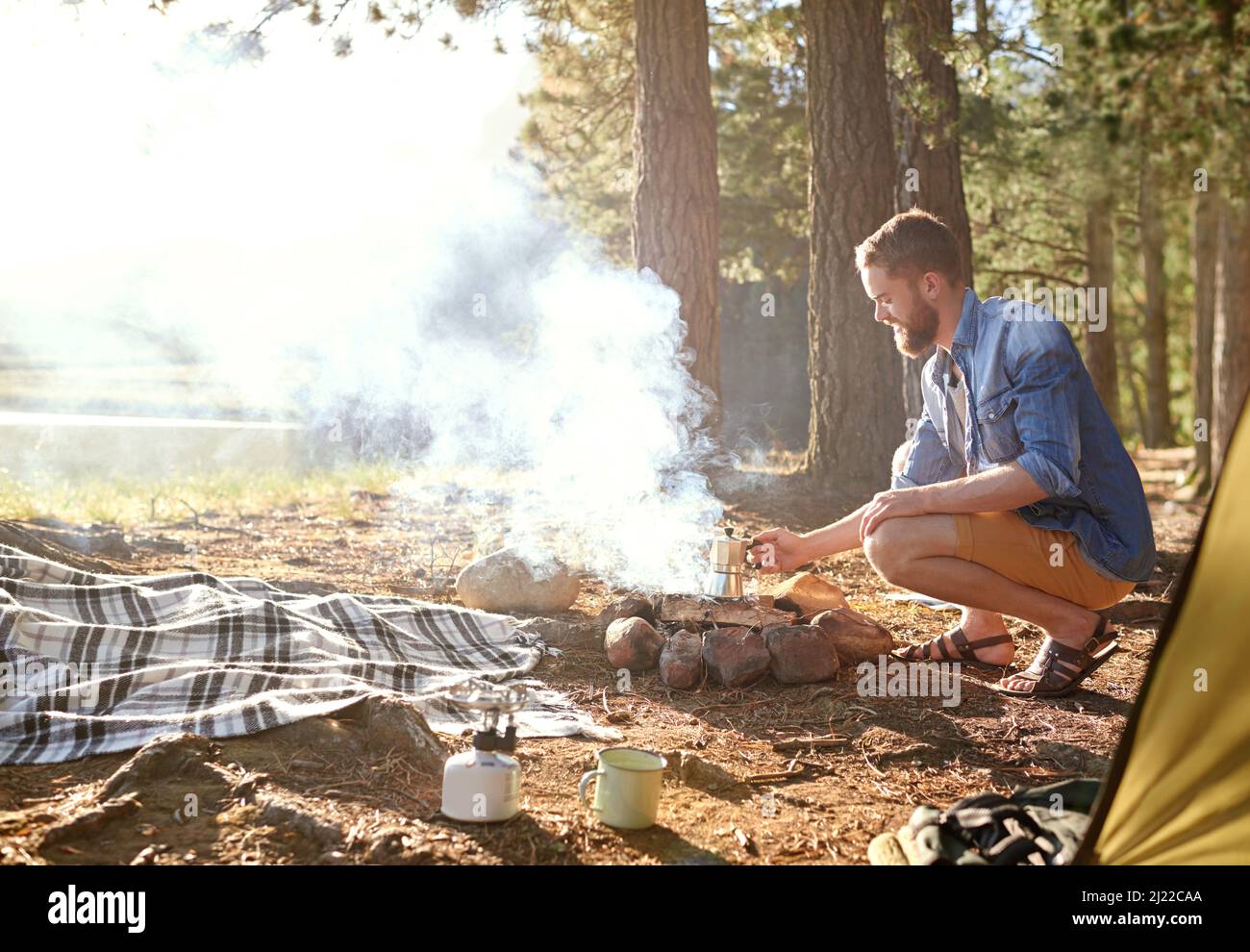Into the woods. Shot of a young man tending to a fire at his campsite ...