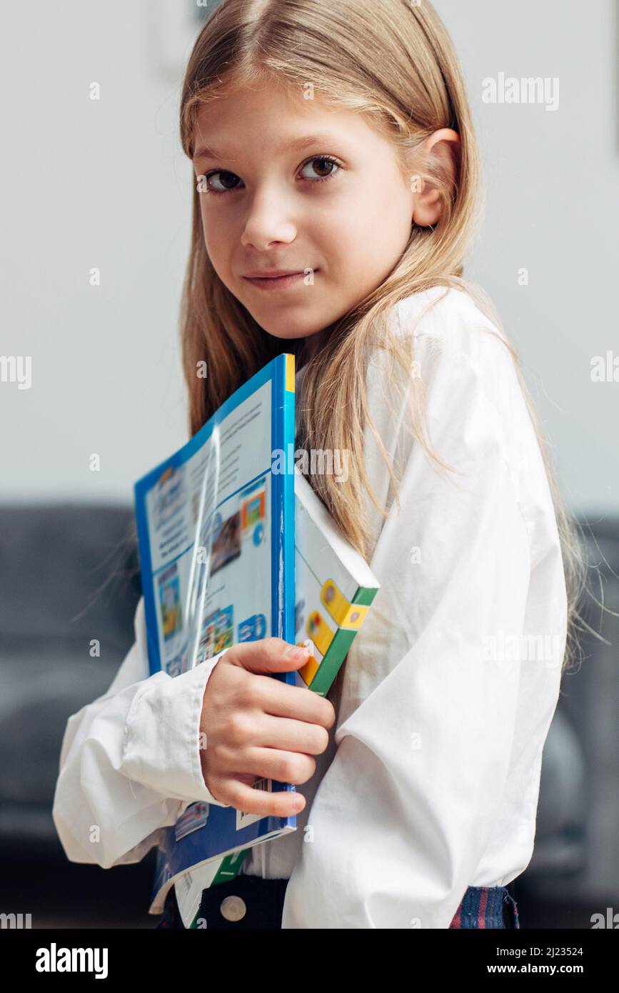 Elementary school girl stands with a textbook and looks at the camera ...