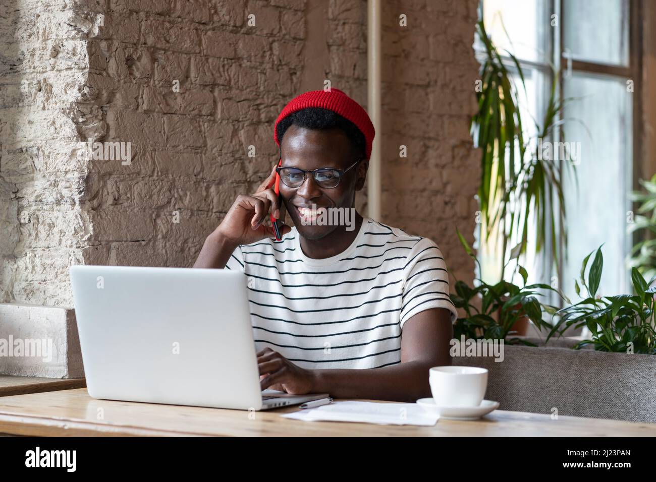 Smiling African freelancer man talking on phone, holding cup of coffee ...