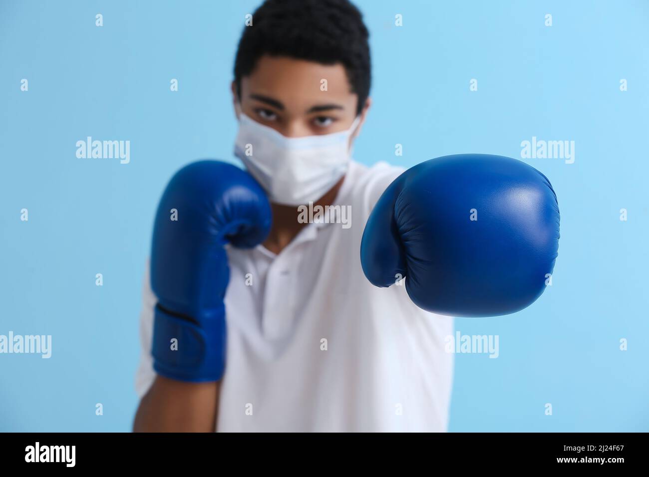 Young African-American man in medical mask with boxing gloves on blue ...
