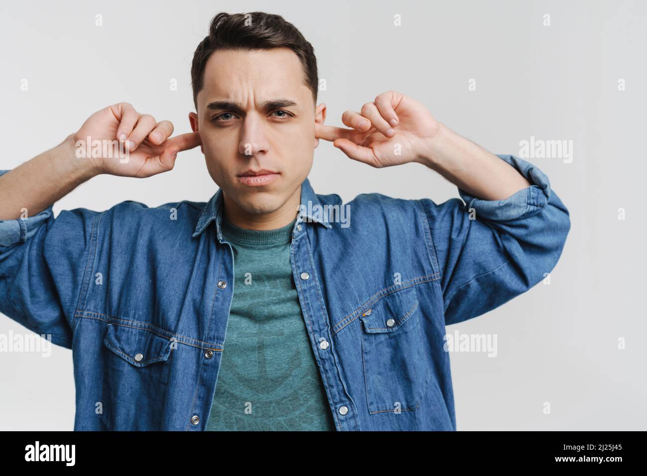 Young brunette man frowning and plugging his ears isolated over white ...