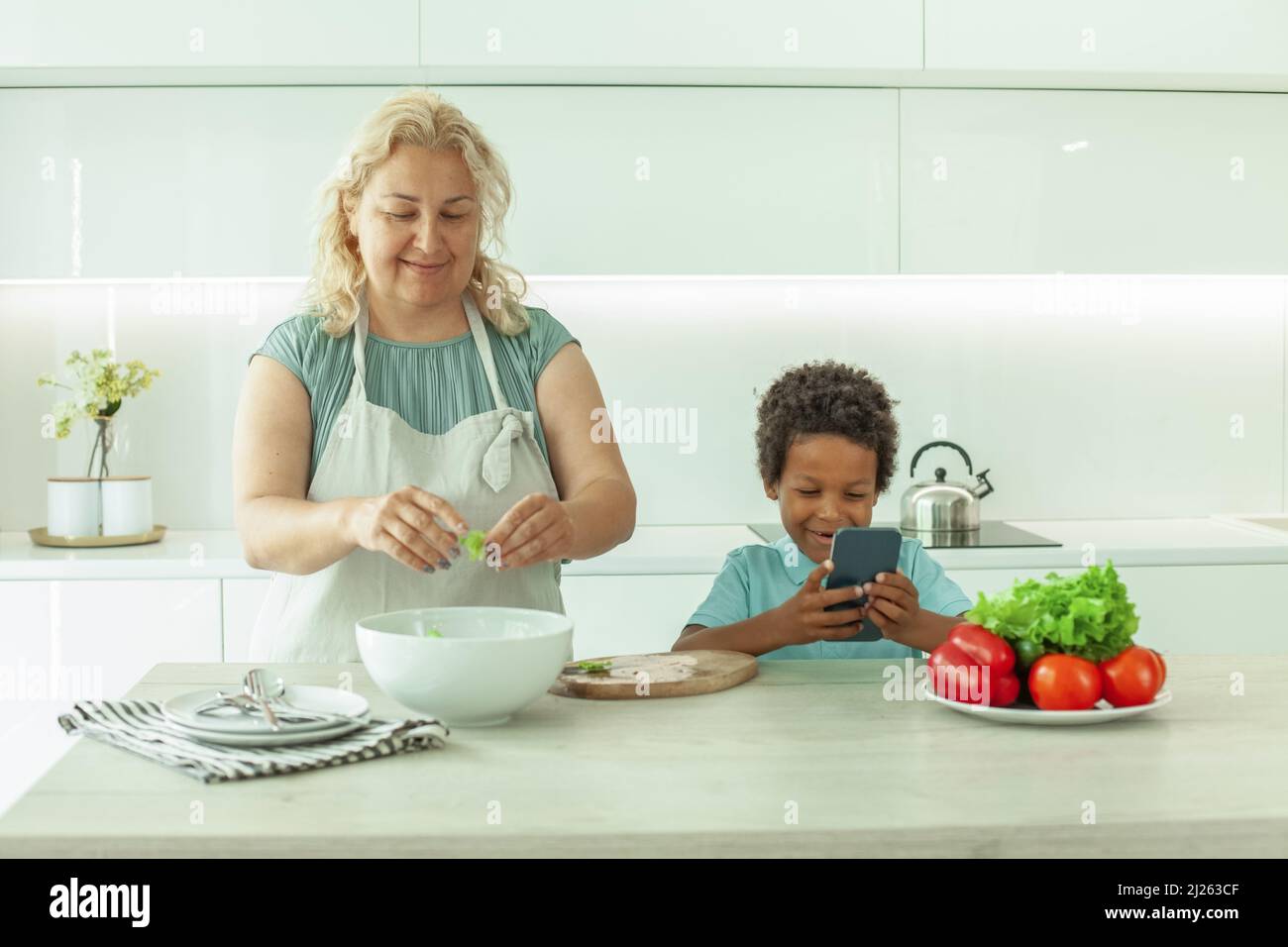 Happy kid boy using phone while cooking in kitchen with her mother ...
