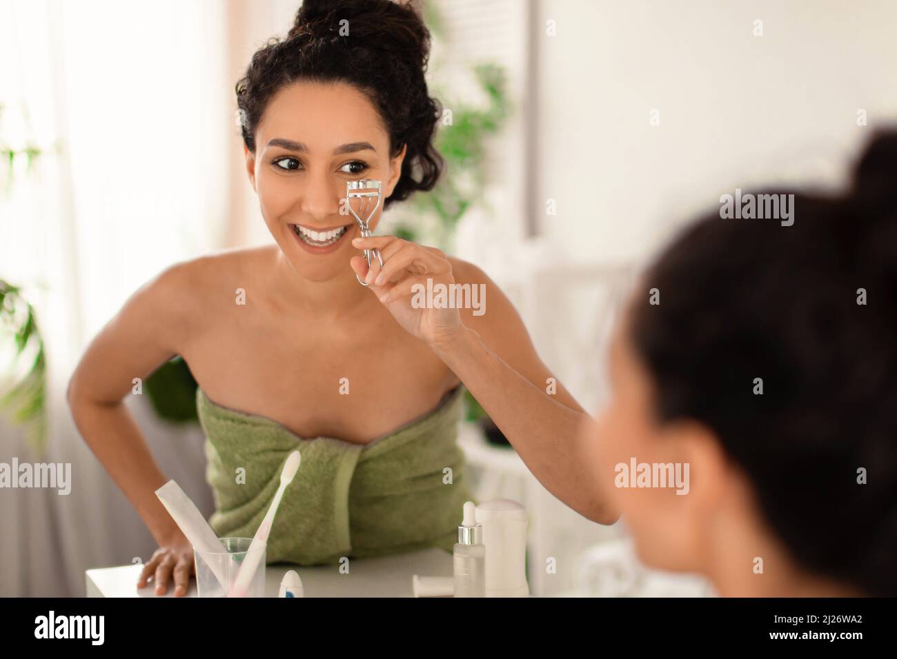 Pretty smiling millennial woman in towel using eyelash curler near ...