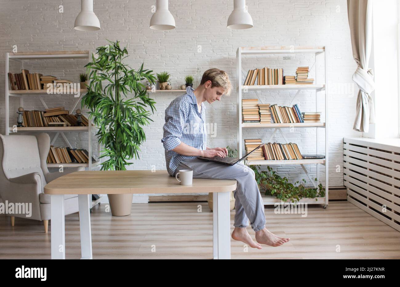 A young programmer guy is sitting on top of a table with his legs ...