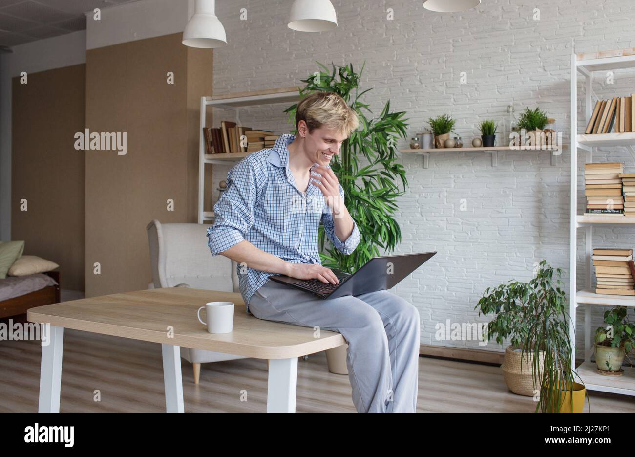 A young programmer guy is sitting on top of a table with his legs ...