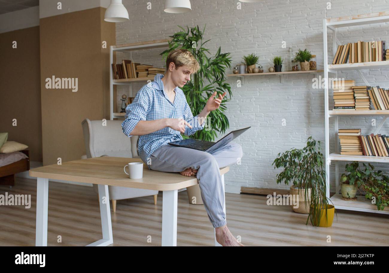 A young programmer guy is sitting on top of a table with his legs ...