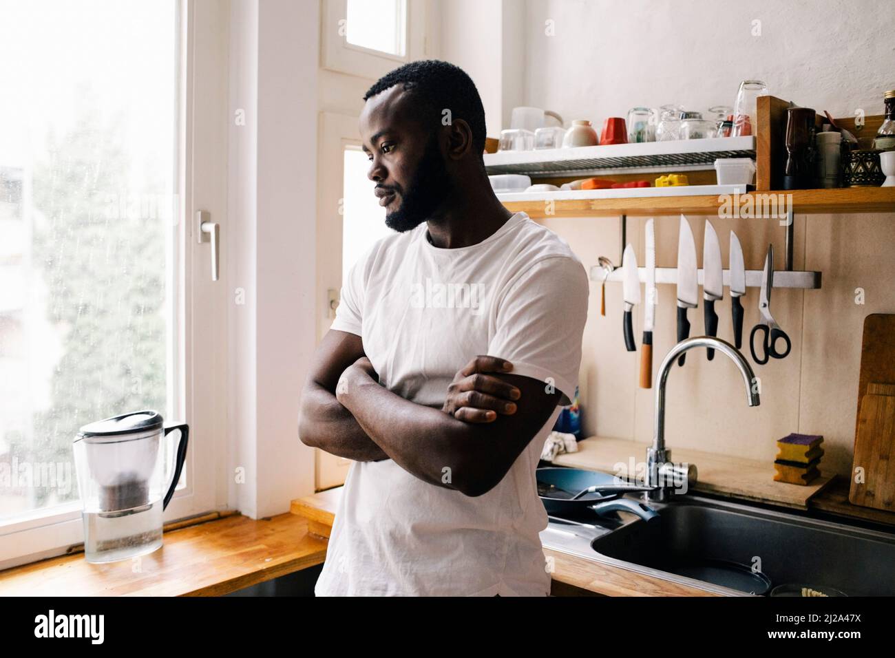 Thoughtful man with arms crossed looking down while leaning on kitchen ...