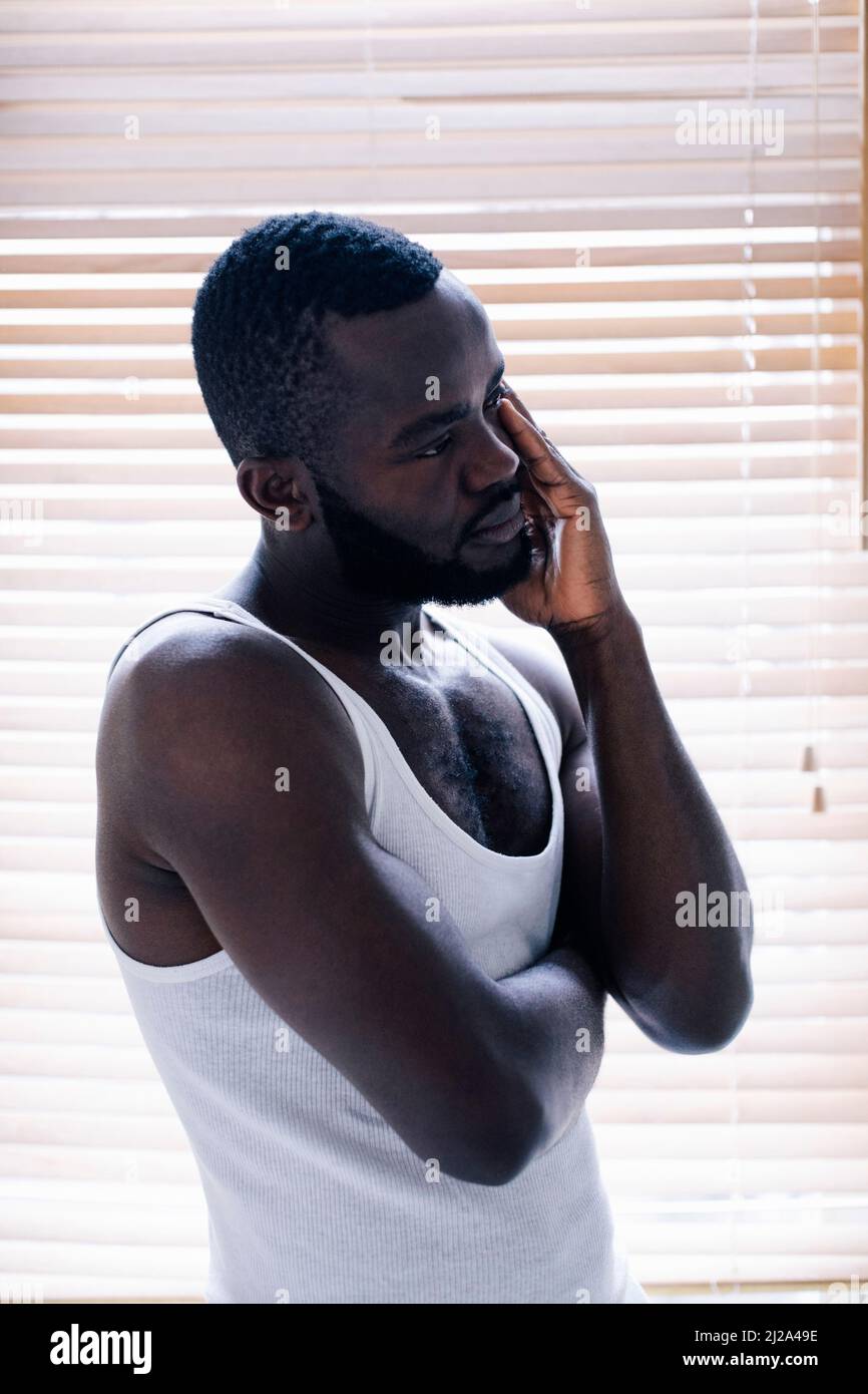 Emotionally disturbed man touching face while standing against blinds ...