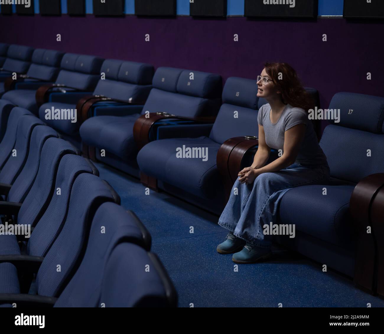 Caucasian red-haired woman sits on the last row in a cinema in an empty hall. The girl is ...
