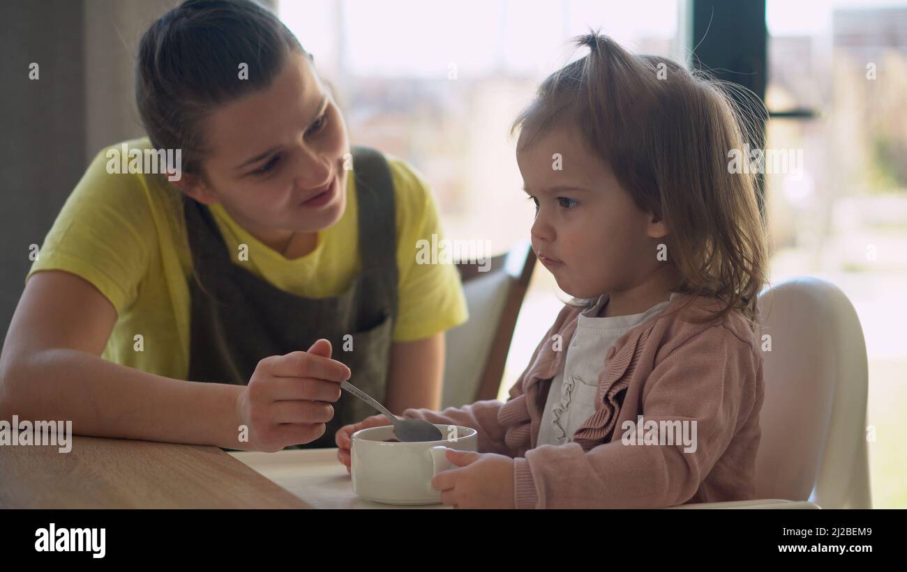 Closeup mom feed young baby in white feeding up high chair, kid is ...