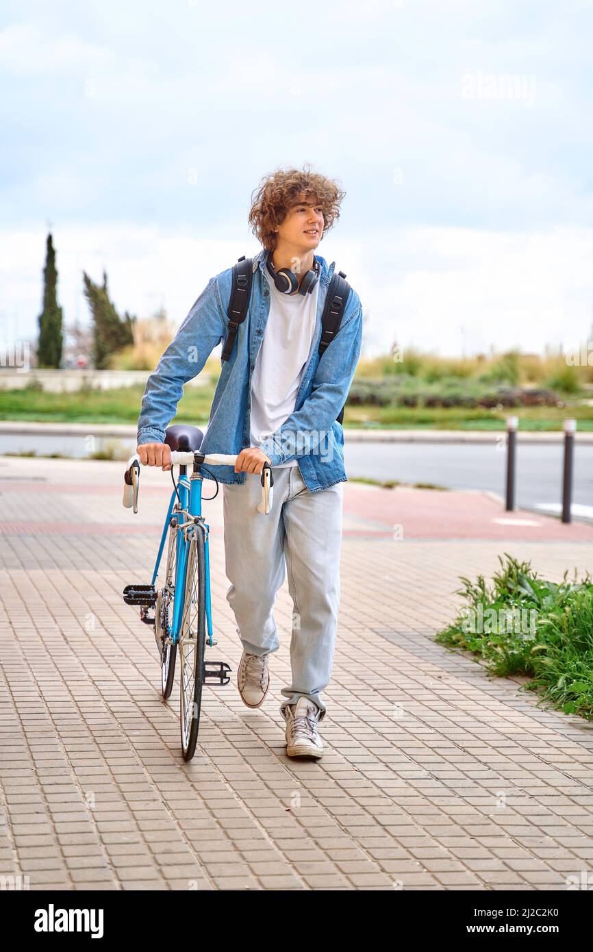 Young curly-haired student with headphones walking to college holding ...