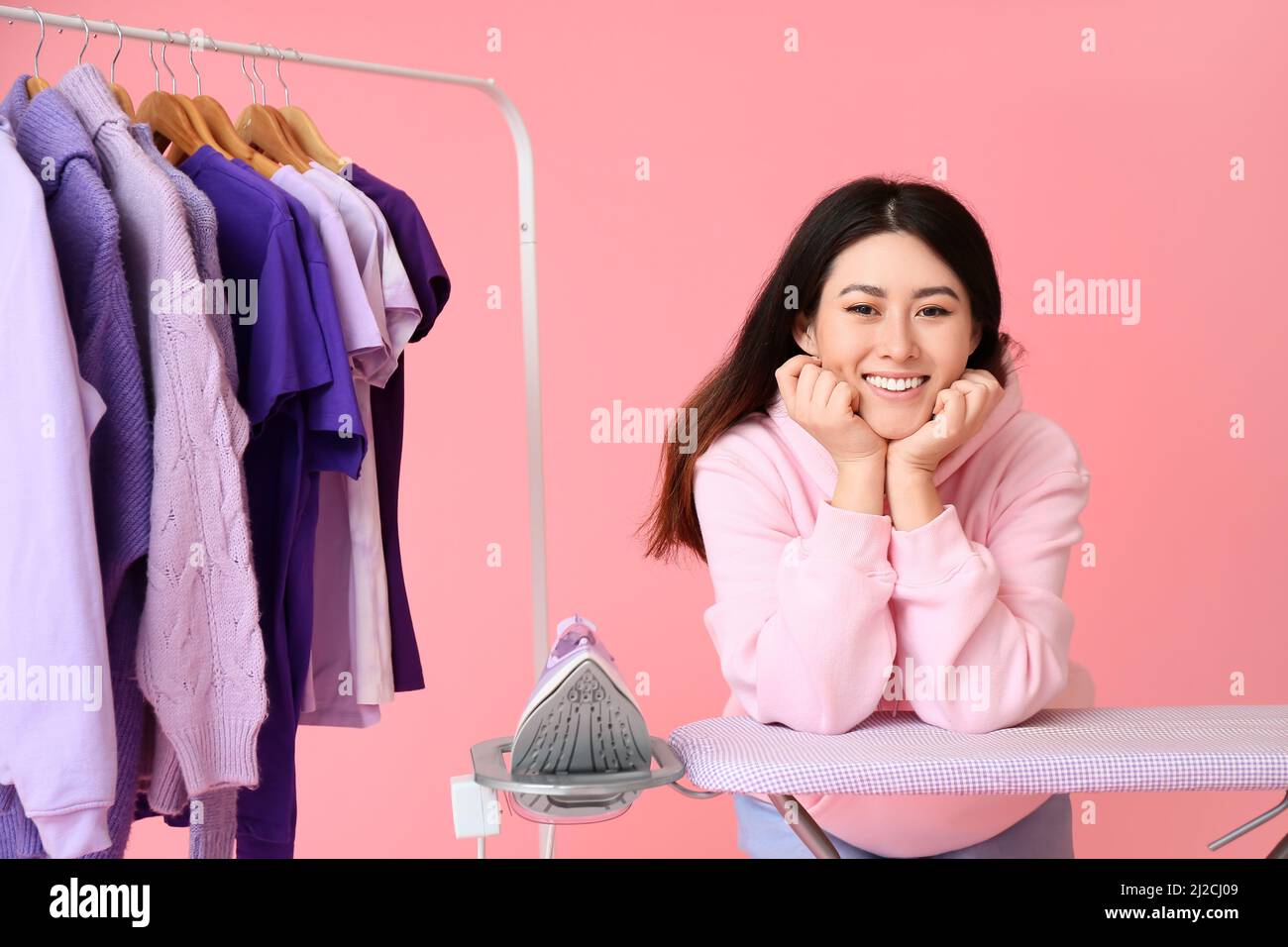 Young Asian woman with rack, clothes and ironing board on pink ...
